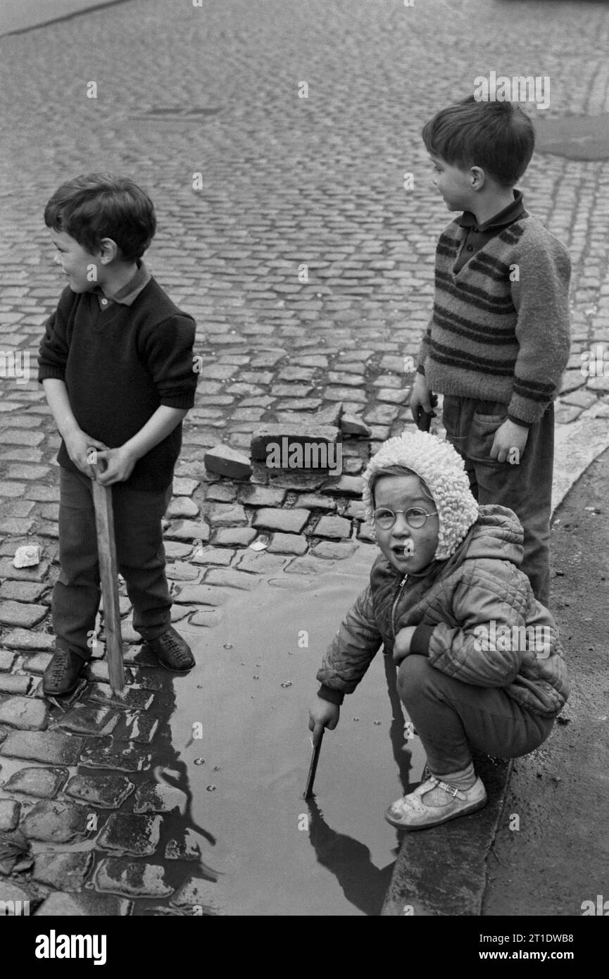 Children playing with sticks in a rain puddle on a cobbled street ...
