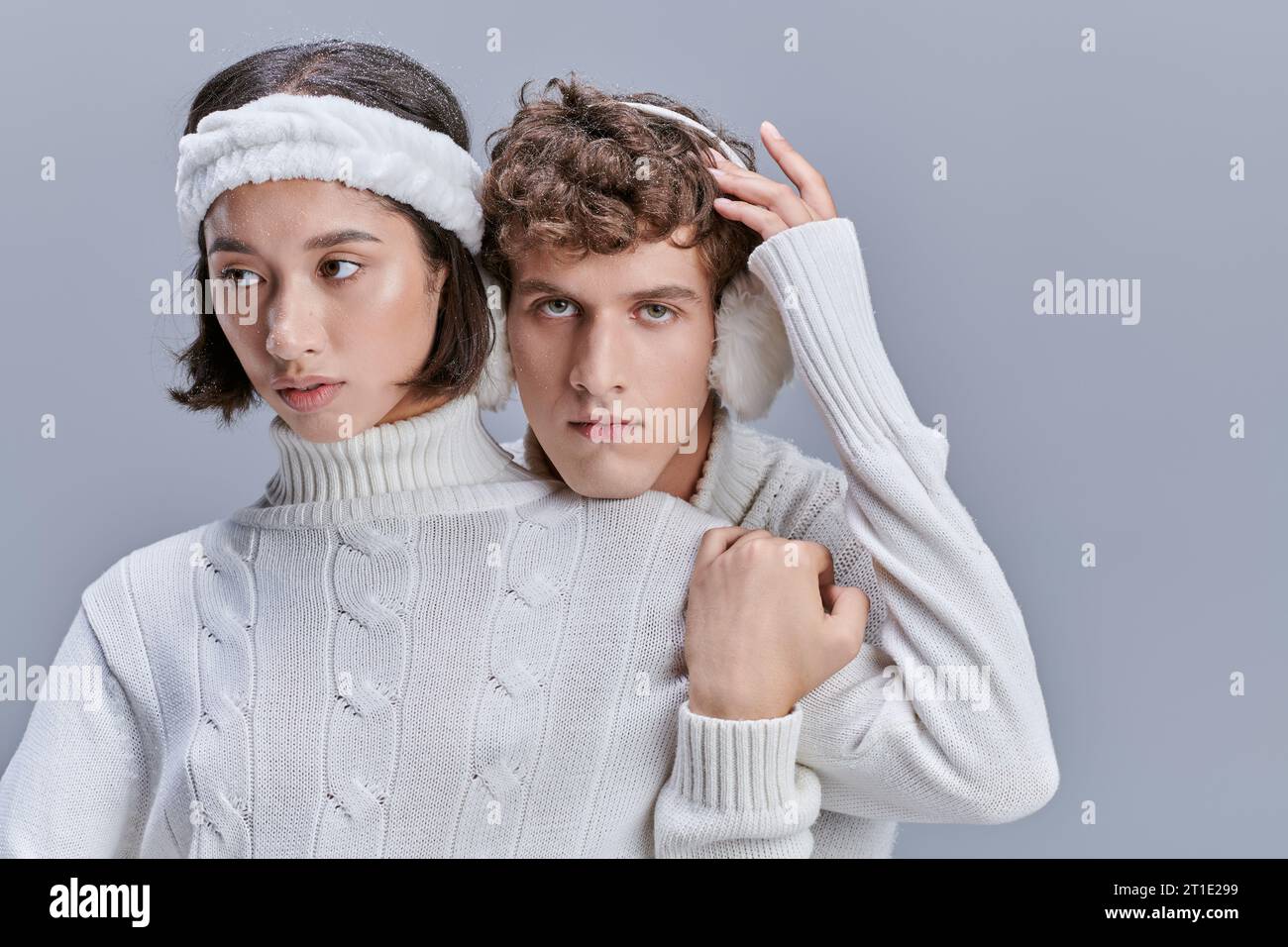 charming woman touching snowy hair of man in warm earmuffs on grey ...
