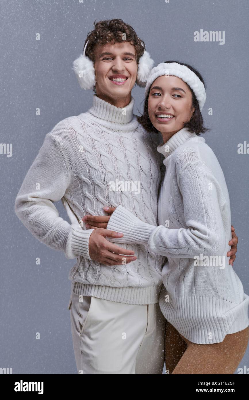 joyful multiethnic couple in knitted sweaters hugging under snowfall on ...