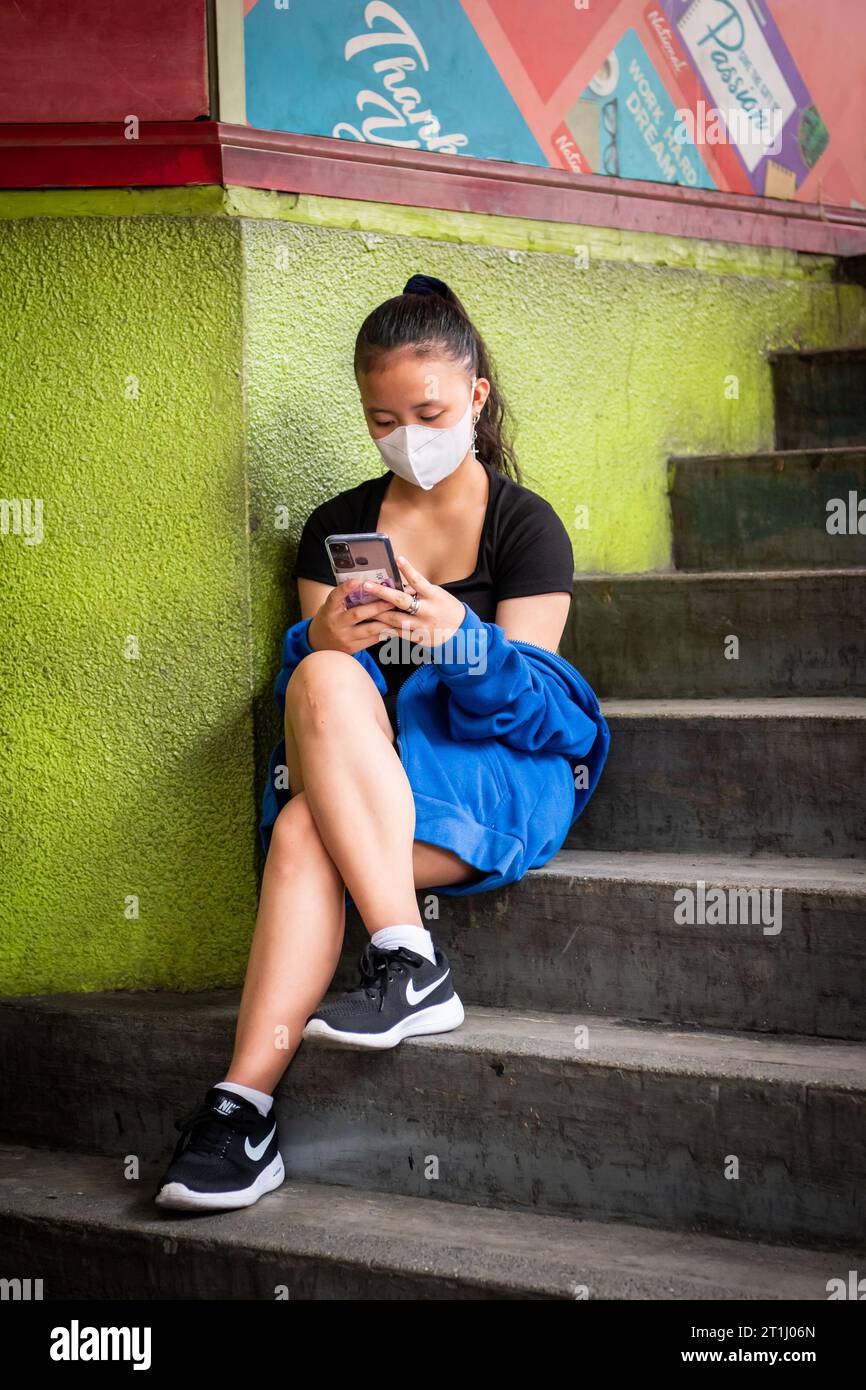 A young Filipino woman checks her mobile phone sat on some steps ...