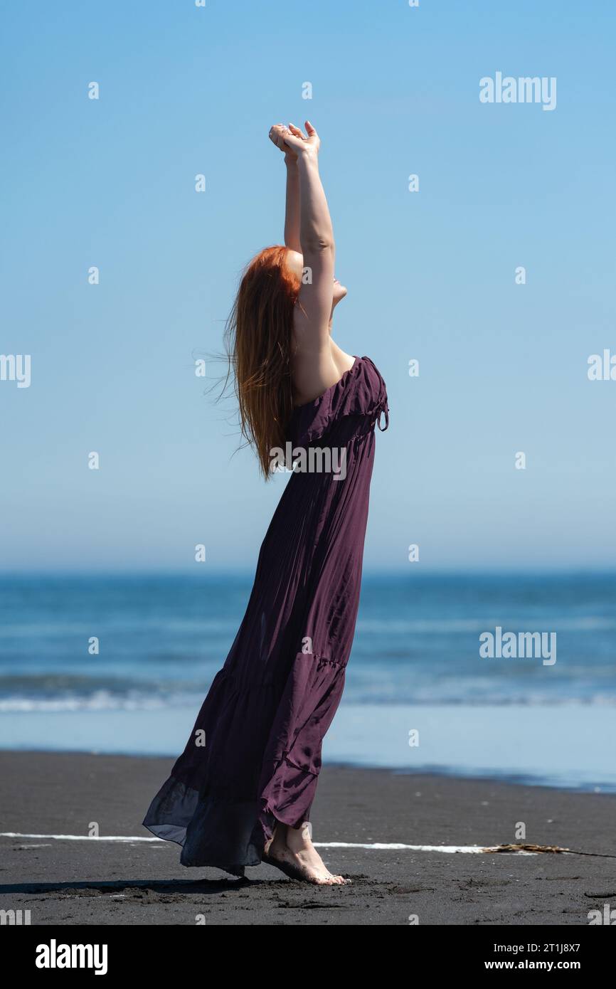 Happy redhead woman in dark puce long dress standing on sandy beach ...