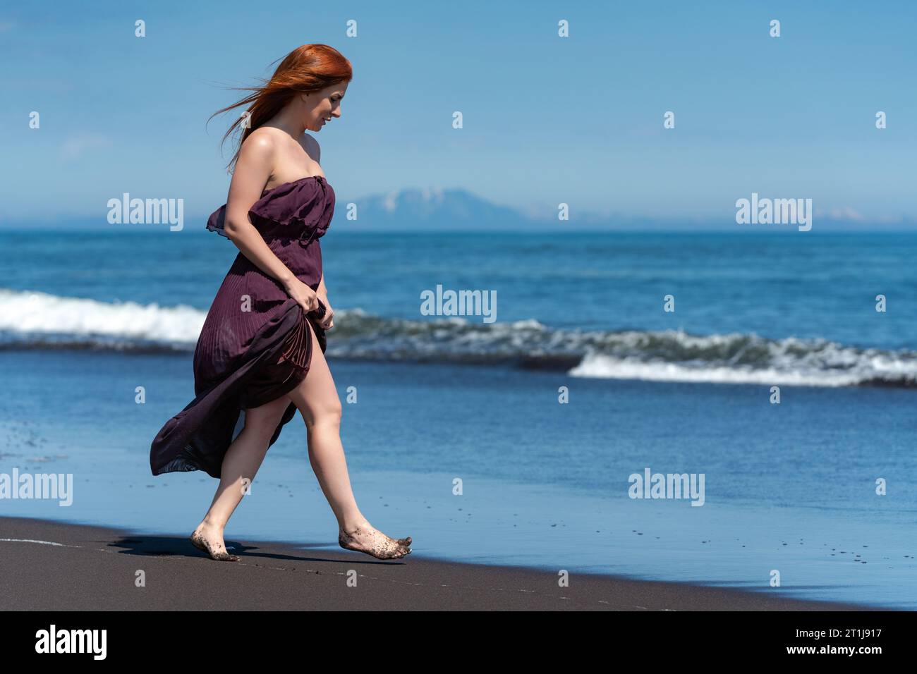 Redhead woman walking along sandy beach in long dress with raised hem ...