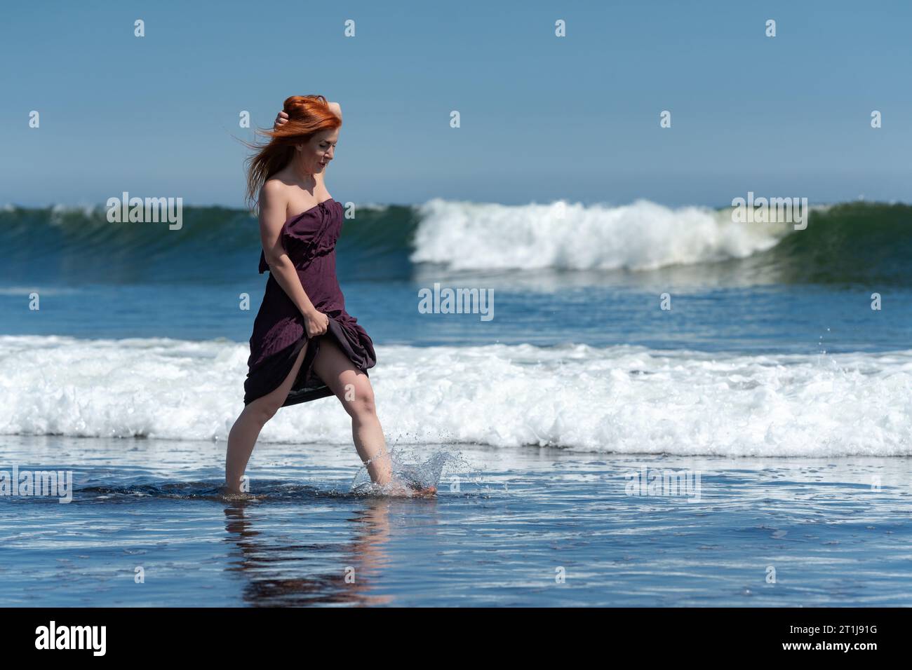 Happy redhead woman walking ankle deep in water of breaking ocean waves ...