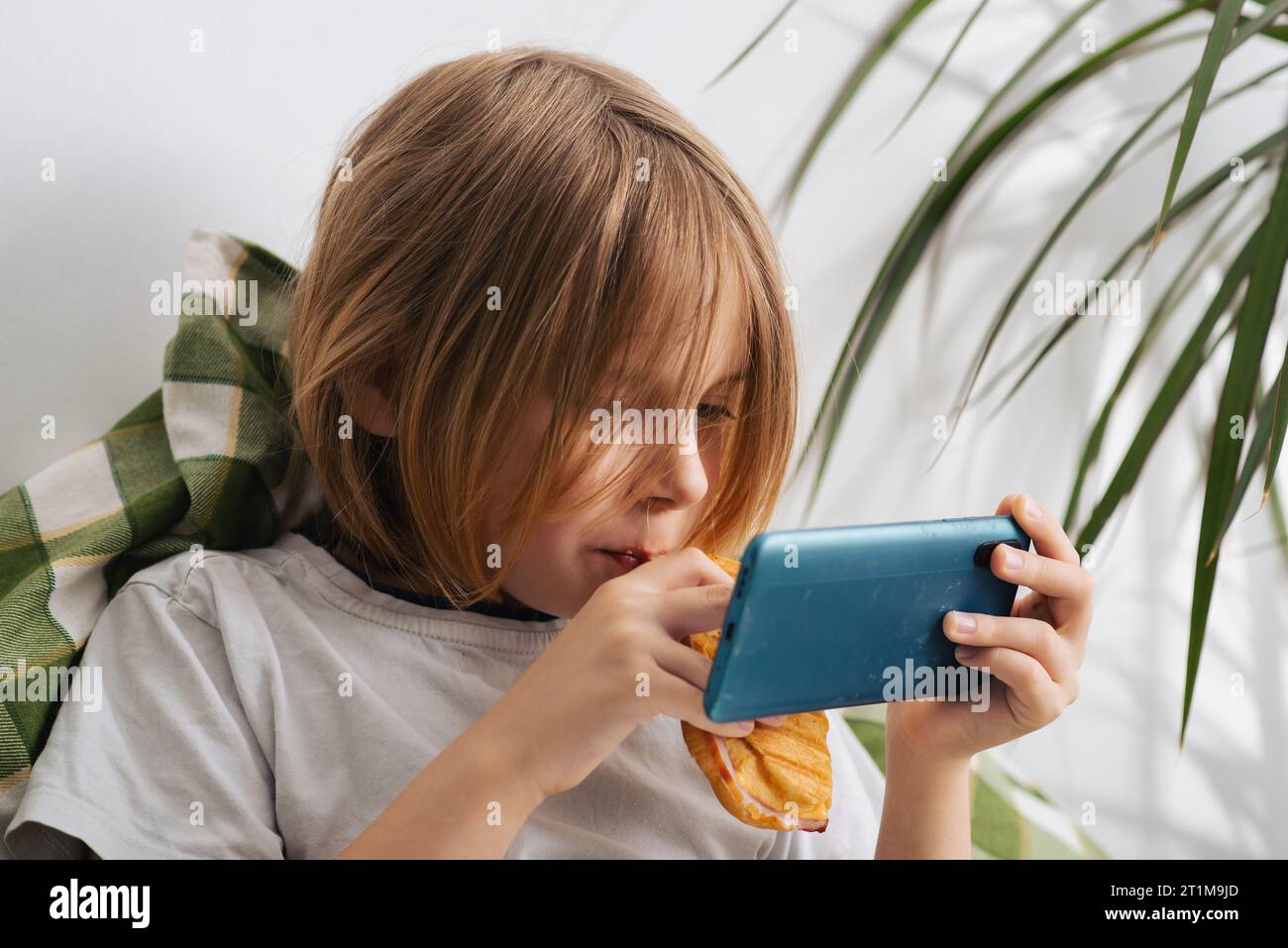 A young boy lies in bed looking at his phone and eating a sandwich ...