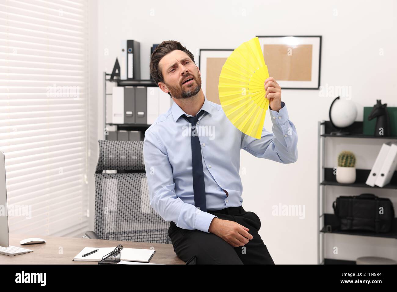 Bearded businessman waving yellow hand fan to cool himself in office ...