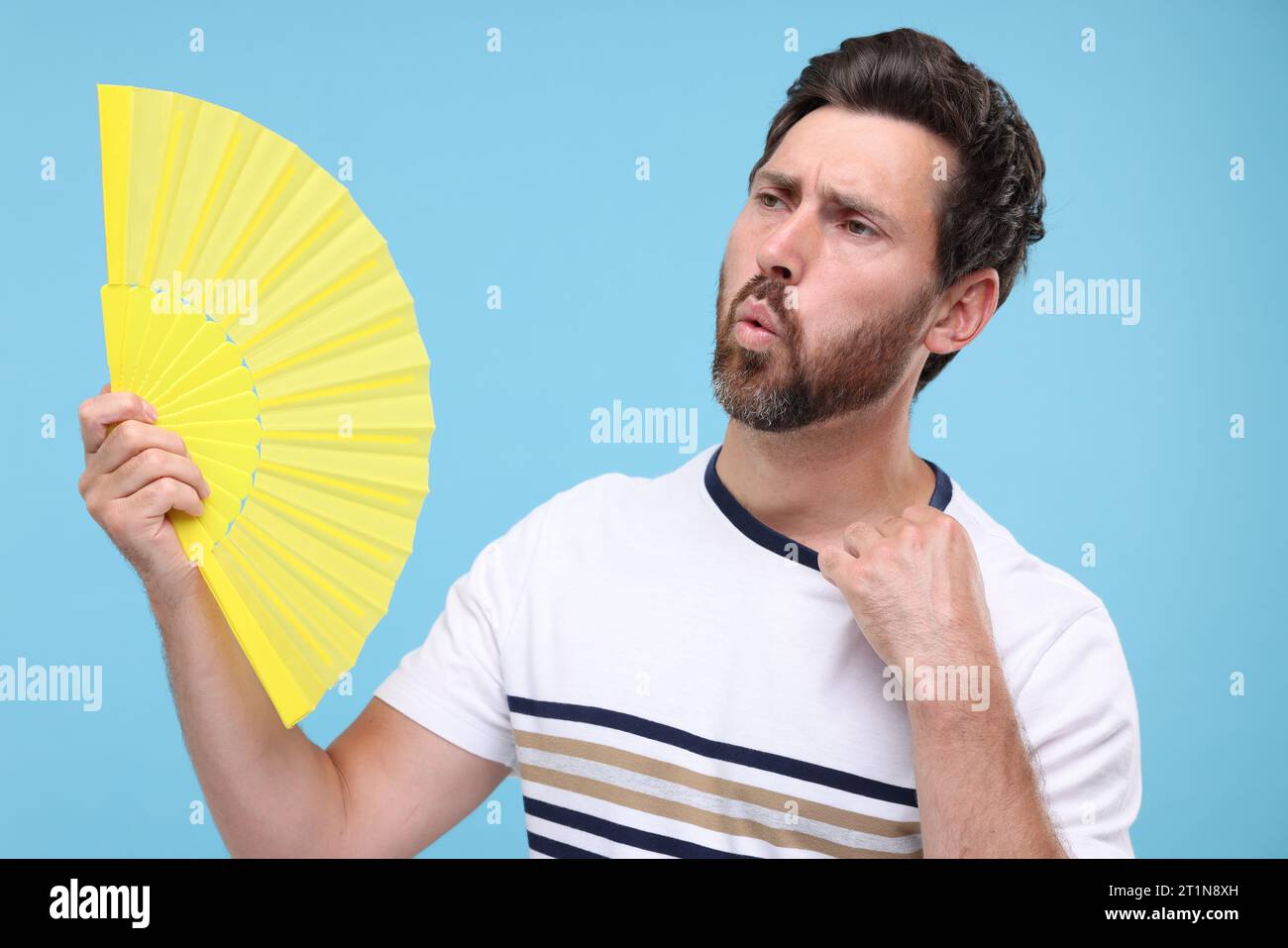 Unhappy man with hand fan suffering from heat on light blue background ...