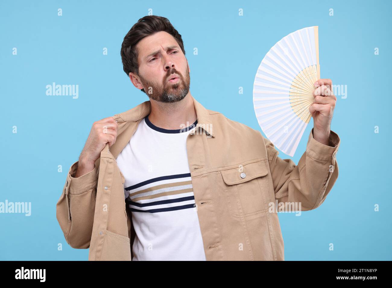 Unhappy man with hand fan suffering from heat on light blue background ...