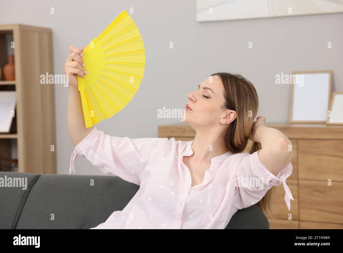 Woman waving yellow hand fan to cool herself on sofa at home Stock ...