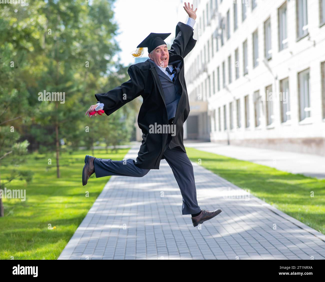 Old happy man in graduation gown jumping outdoors and holding diploma ...