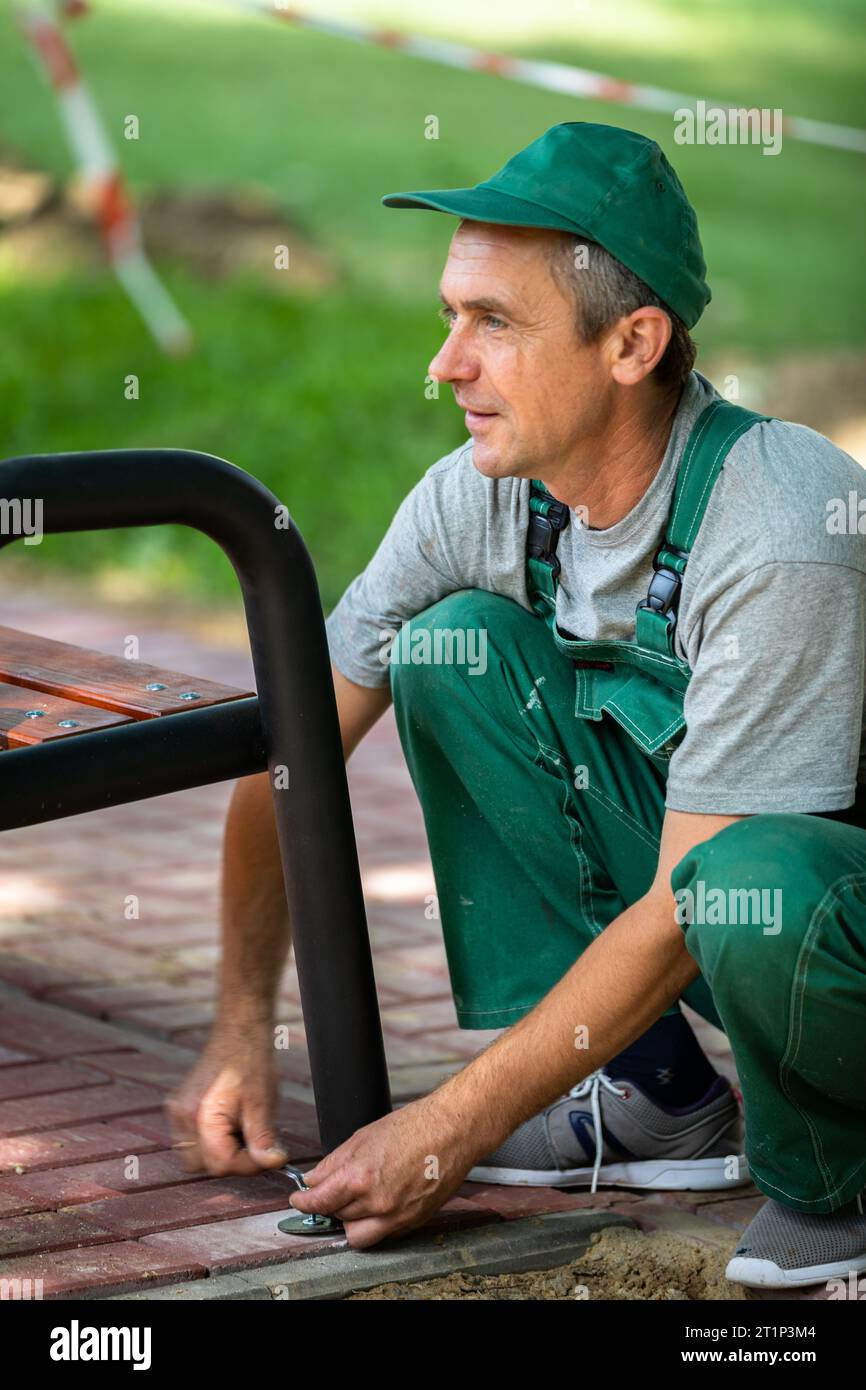 A construction worker, squatting and screwing a park bench to the ...