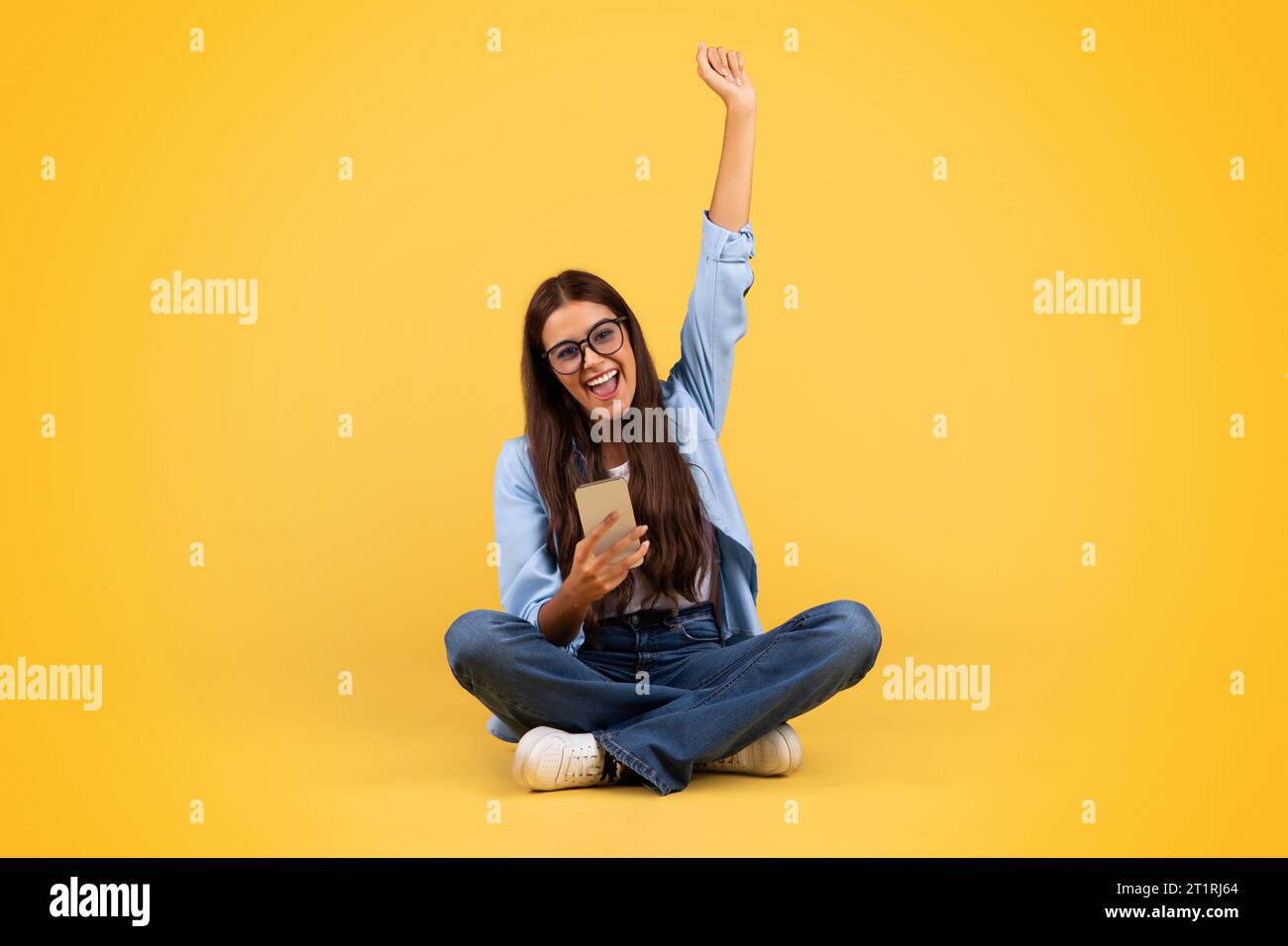 Satisfied teen student woman in glasses look at phone, raise hand up ...