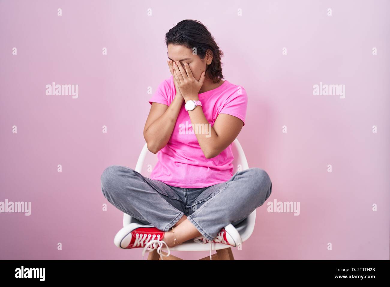 Hispanic young woman sitting on chair over pink background with sad ...