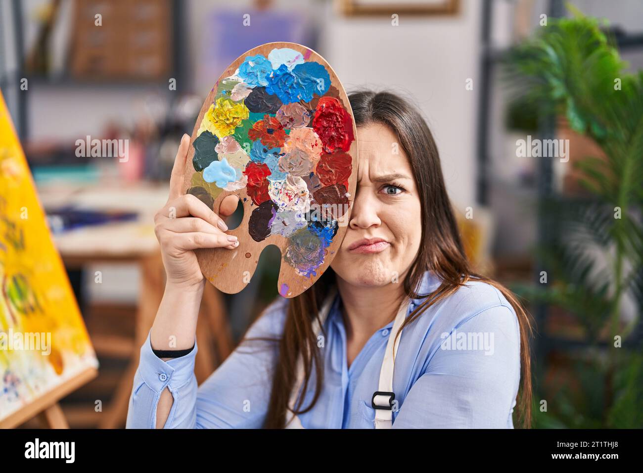Young brunette woman covering face with painter palette skeptic and ...