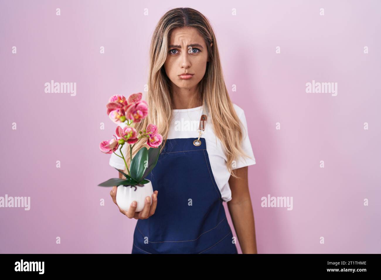 Young blonde woman wearing gardener apron holding plant depressed and ...