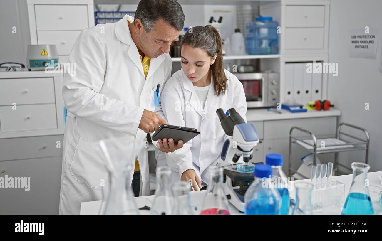 Man and woman, two focused scientists, masterfully manipulate a microscope and touchpad in a ...