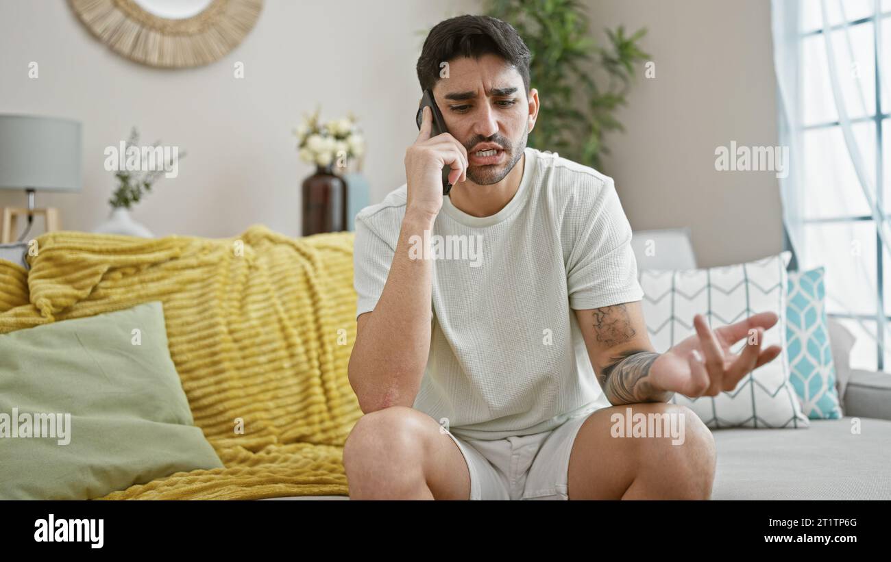 Young hispanic man arguing by smartphone sitting on sofa angry at home ...