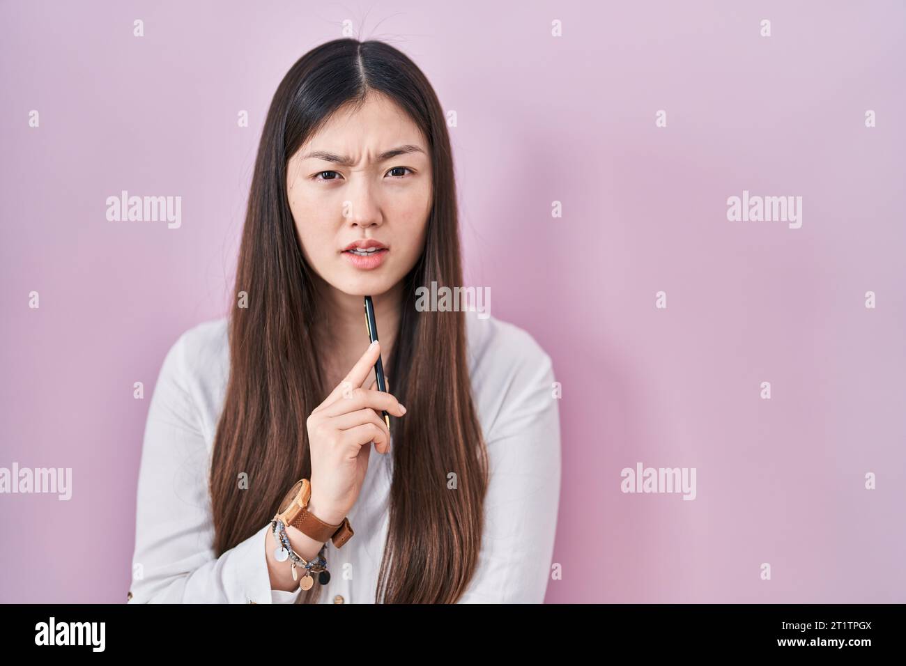 Chinese young woman holding pencil over pink background clueless and ...