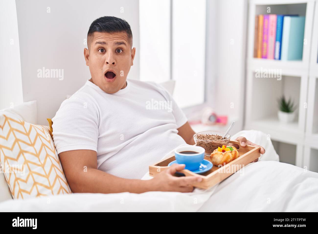 Young hispanic man eating breakfast in the bed scared and amazed with ...