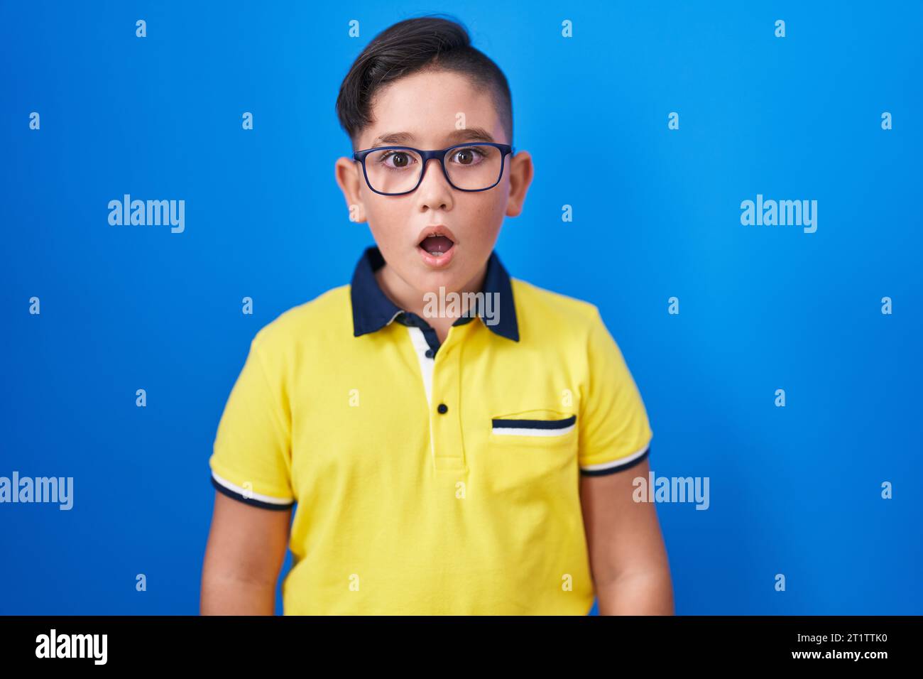 Young hispanic kid standing over blue background scared and amazed with ...