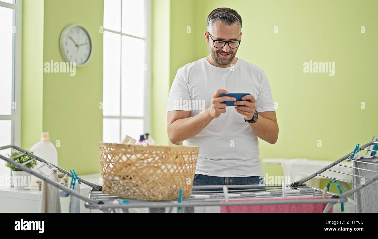 Grey-haired man playing video game standing by clothesline at laundry ...