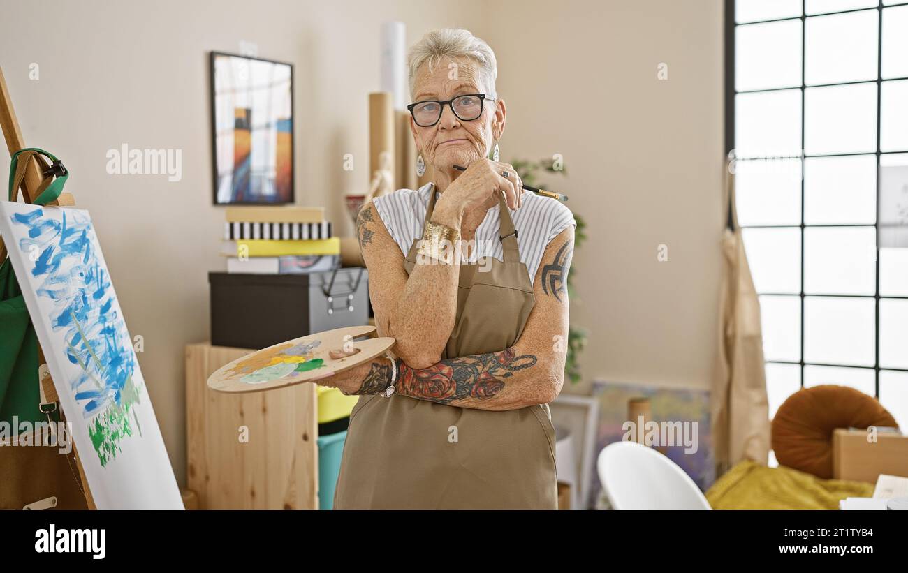 Grey-haired senior woman artist, apron-clad and glasses perched, stands ...