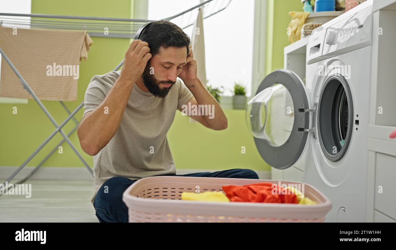 Young hispanic man listening to music washing clothes at laundry room ...