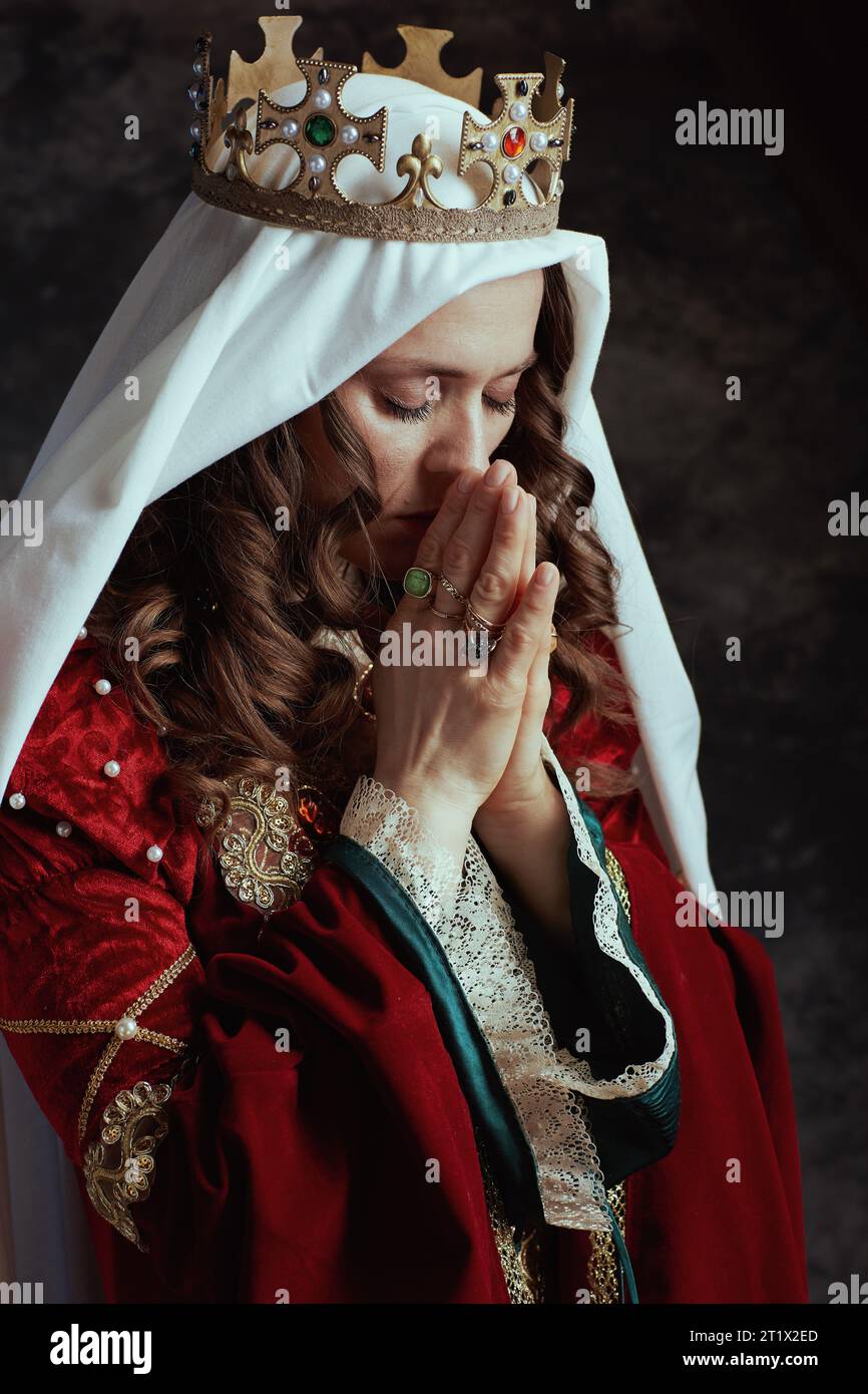 medieval queen in red dress with veil and crown praying on dark gray ...
