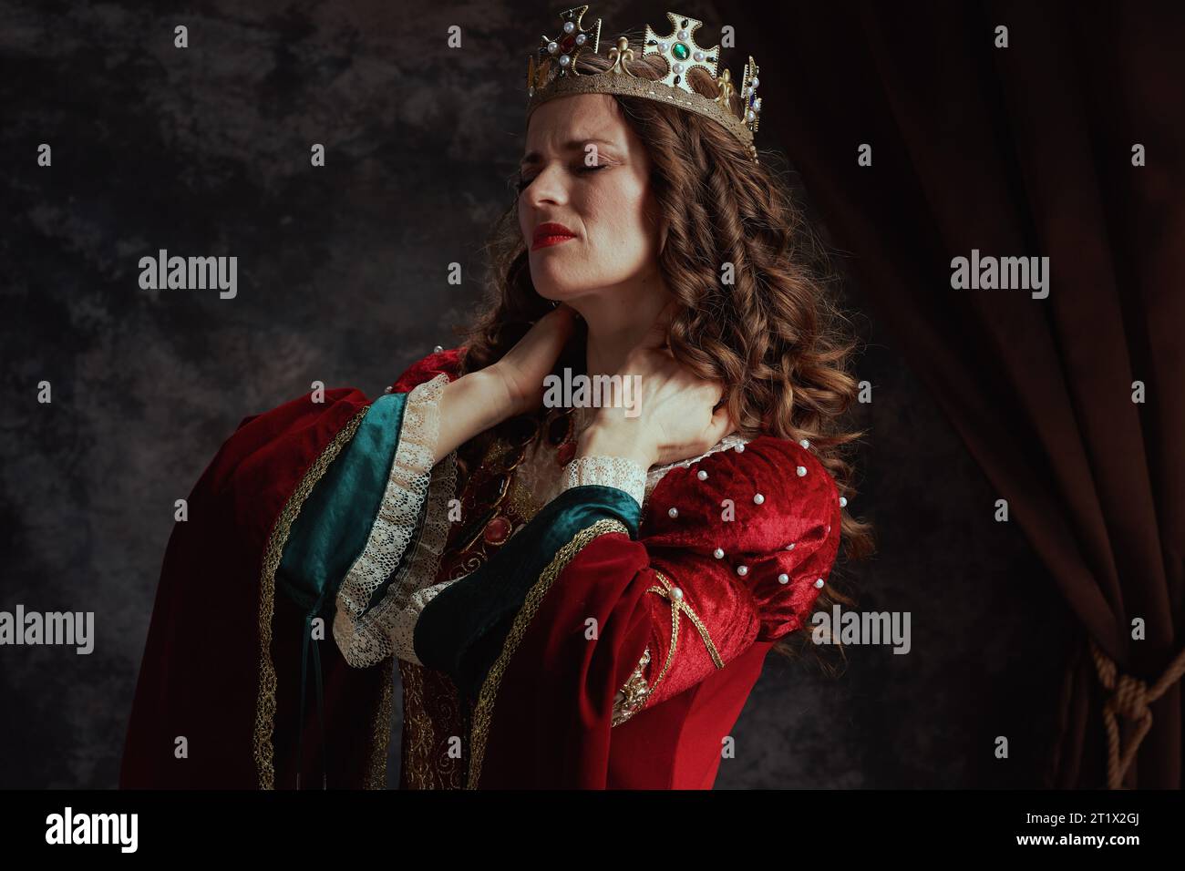 tired medieval queen in red dress with crown having neck pain on dark ...