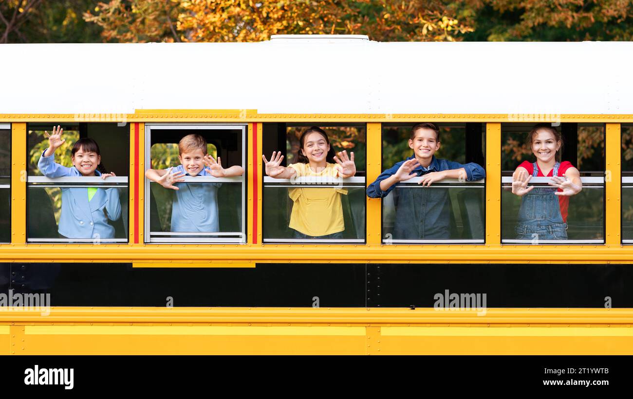 Joyful kids looking out from yellow school bus, waving hands at camera ...
