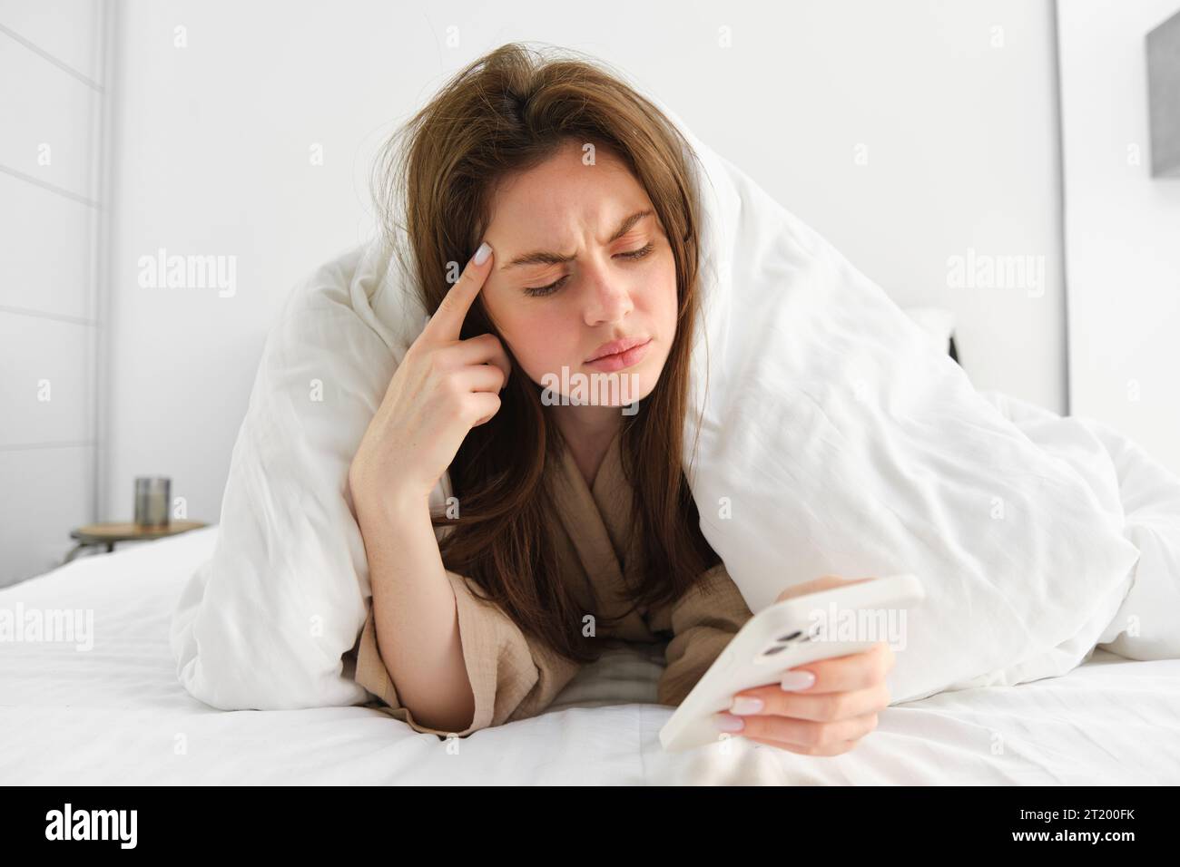 Woman lying in bed in morning, looking confused at smartphone, reading ...