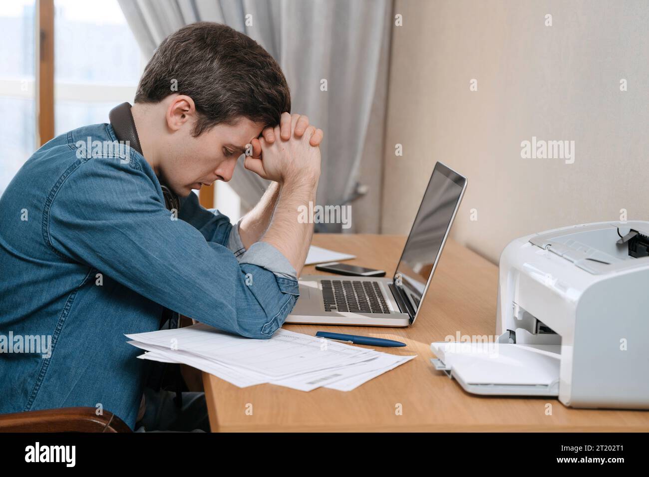 Confused tired young man sitting at his desk with laptop, hands folded ...