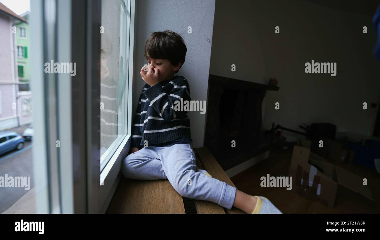 Child nose picking while sitting by window at home looking out from ...