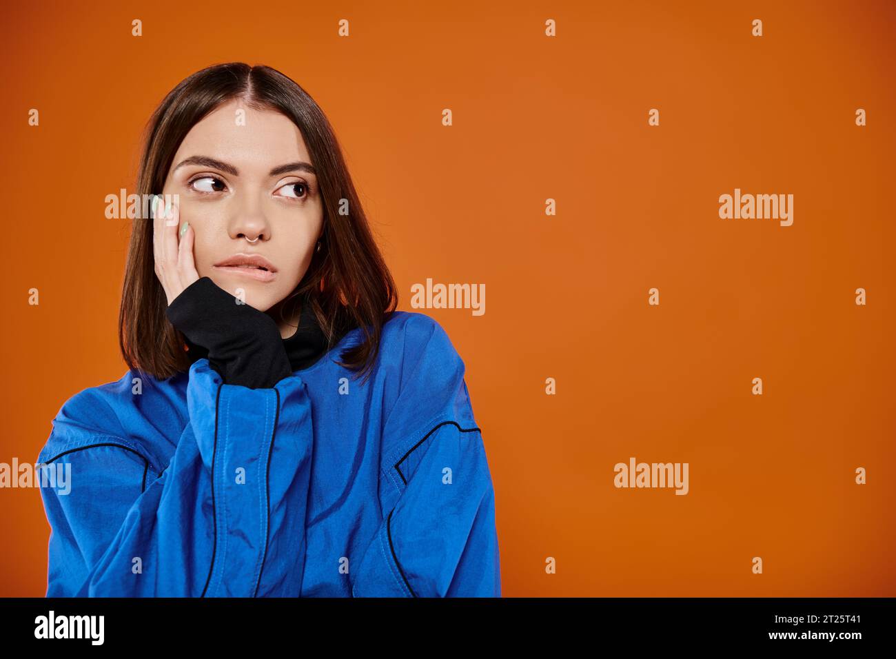 pensive woman with pierced nose biting lip and looking away on orange ...
