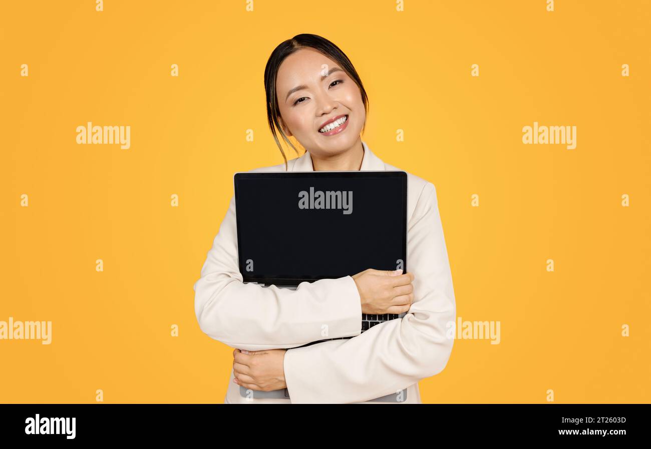 Cheerful korean millennial woman in suit hugs laptop with blank screen ...