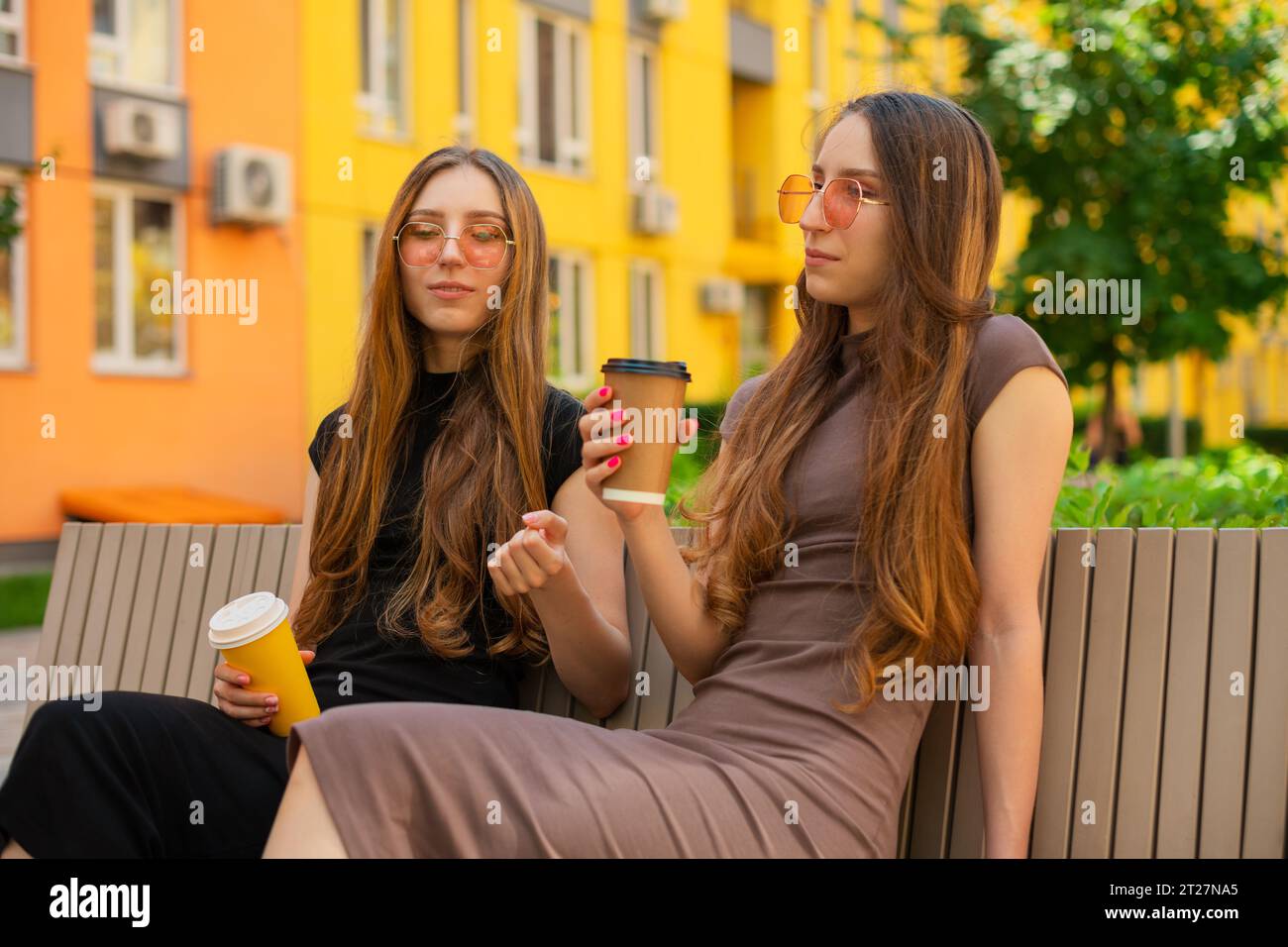 Cute young twin sisters Gen Z with sunglasses sitting on a bench and ...