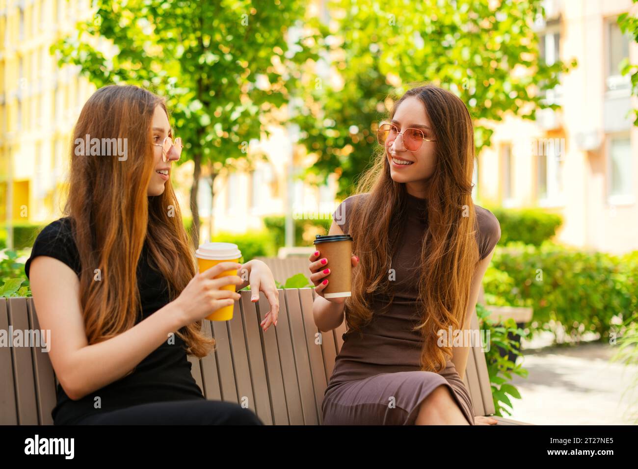 Cute young twin sisters Gen Z with sunglasses sitting on a bench and ...
