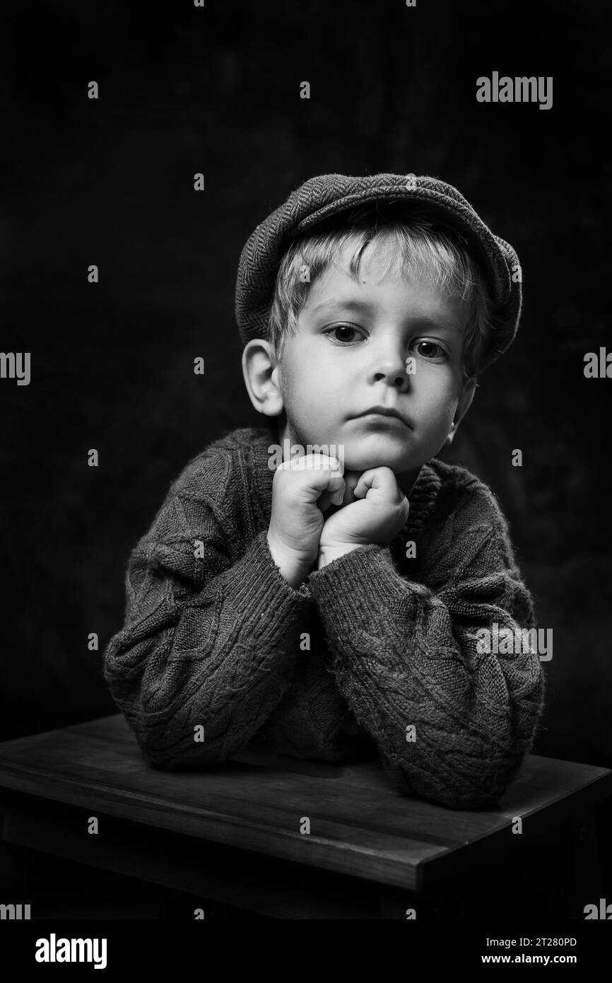 A little boy in a sweater and a retro-style cap on a dark background ...