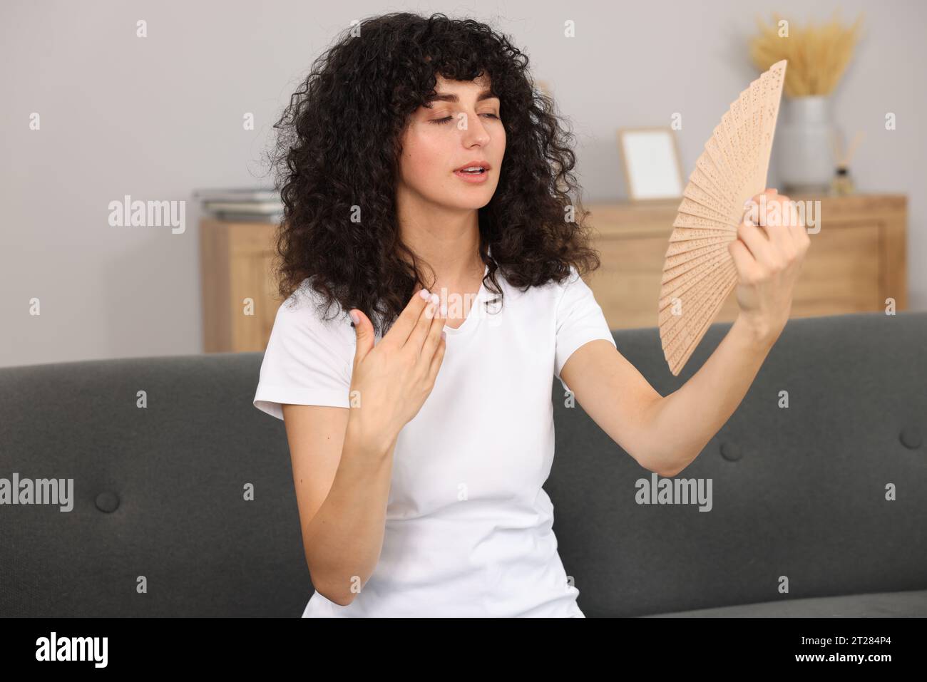 Young woman waving hand fan to cool herself on sofa at home Stock Photo ...
