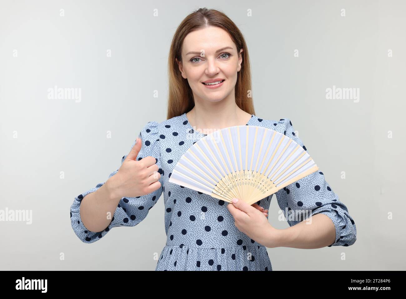 Happy woman with hand fan showing thumb up on light grey background ...