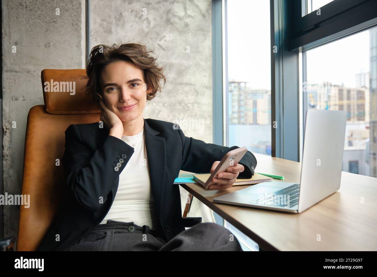 Portrait of corporate woman, saleswoman in an office, using smartphone and laptop, smiling while ...