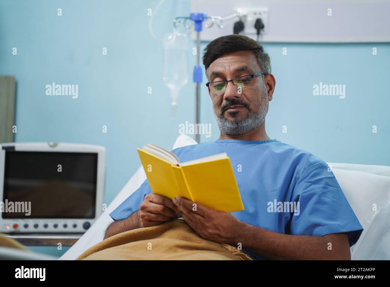 Wide shot Indian elderly senior man reading book at hospital bed after ...
