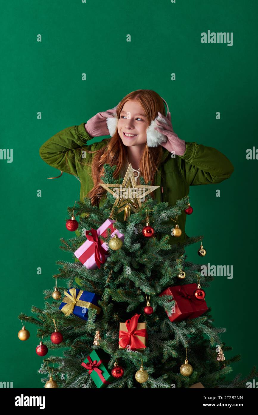 holidays, cheerful girl wearing ear muffs and standing behind decorated ...