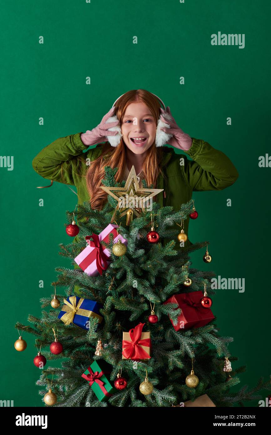 holidays, excited girl wearing ear muffs and standing behind decorated ...
