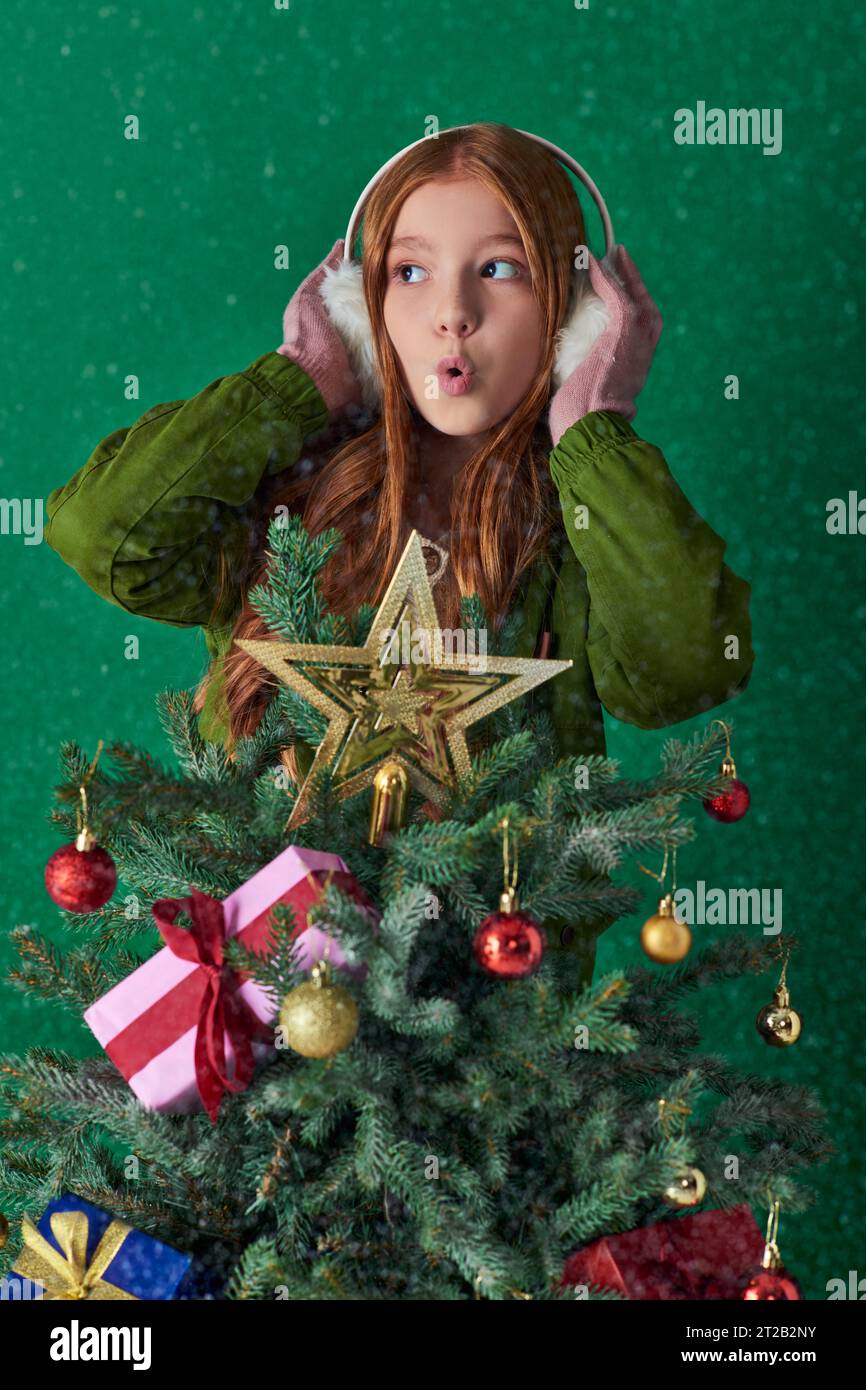 holiday spirit, surprised girl in ear muffs hugging decorated Christmas ...