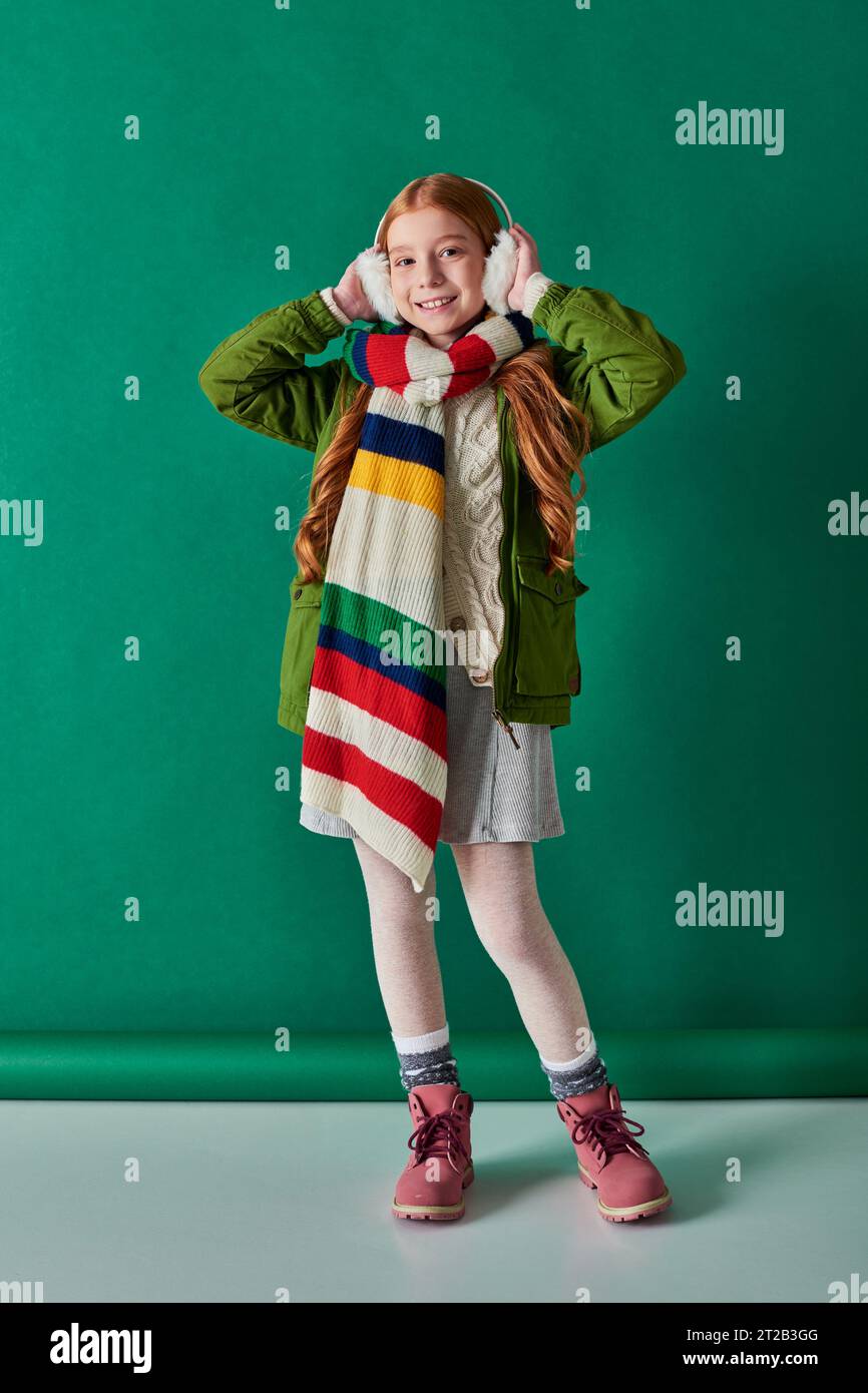 cozy layers, cheerful girl in ear muffs and striped scarf standing in ...