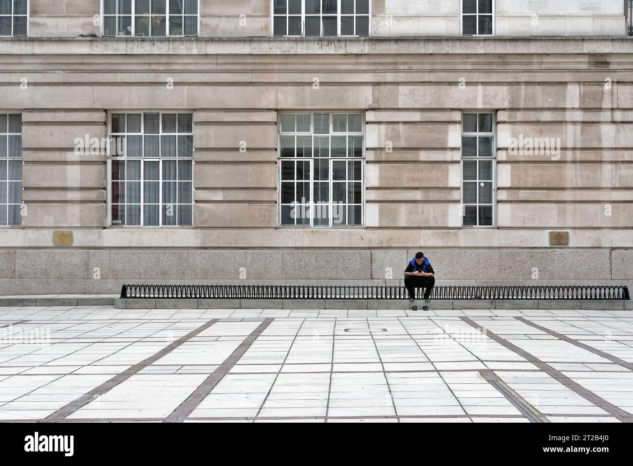 A young man seated on modern seating using a mobile phone in a bleak ...