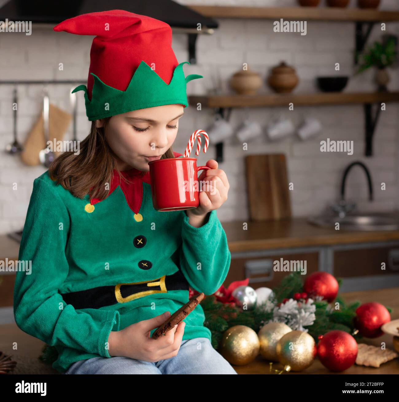 Child in elf costume holding chocolate biscuit and mug with candy cane ...