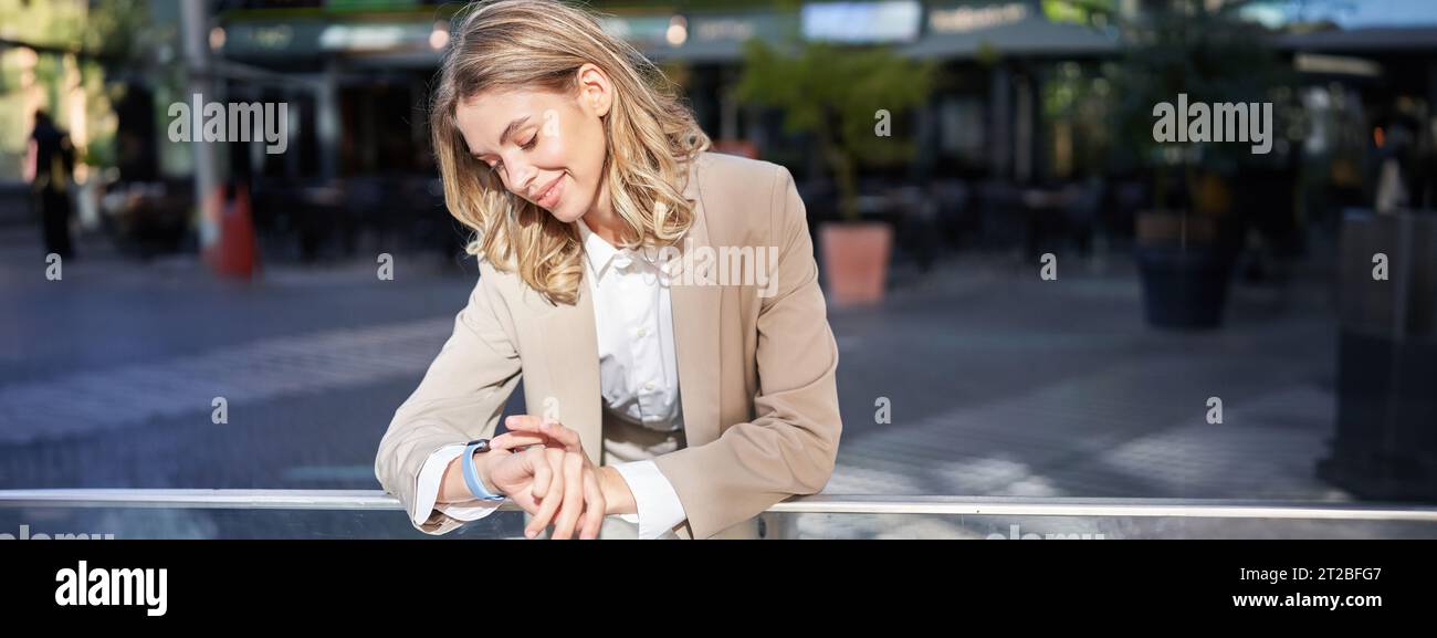 Portrait of corporate woman checks her time, stands on street outside ...