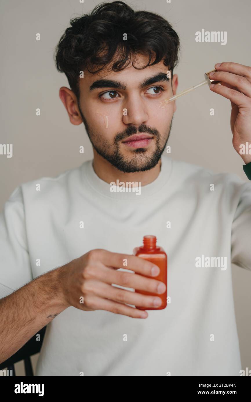 Front view of a young man dropping essential oil or serum on his face ...