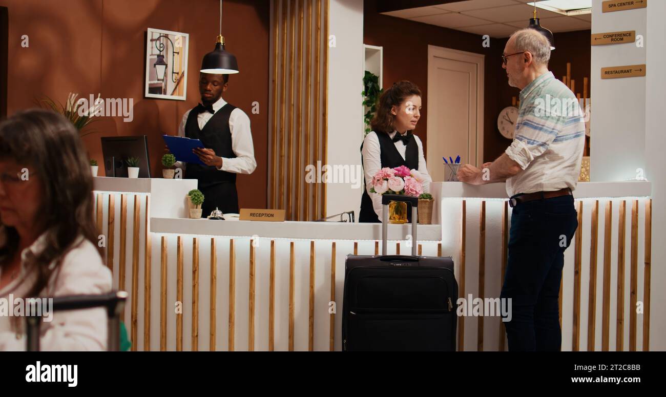 Senior man entering hotel lobby with luggage, asking for concierge services to check in ...